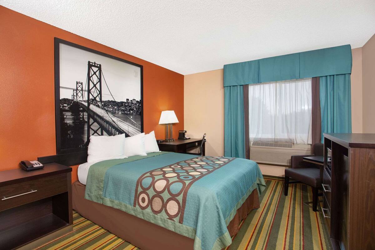 Quarto de hotel com cama, escrivaninha, cadeira, abajur, janela com cortinas e um quadro de uma vista urbana na parede.