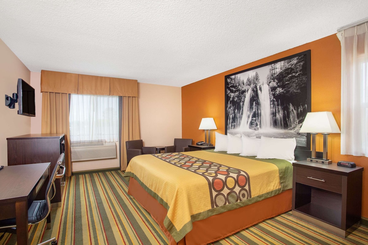 Quarto de hotel com cama, escrivaninha, cadeira, televisão e uma grande janela com cortinas.