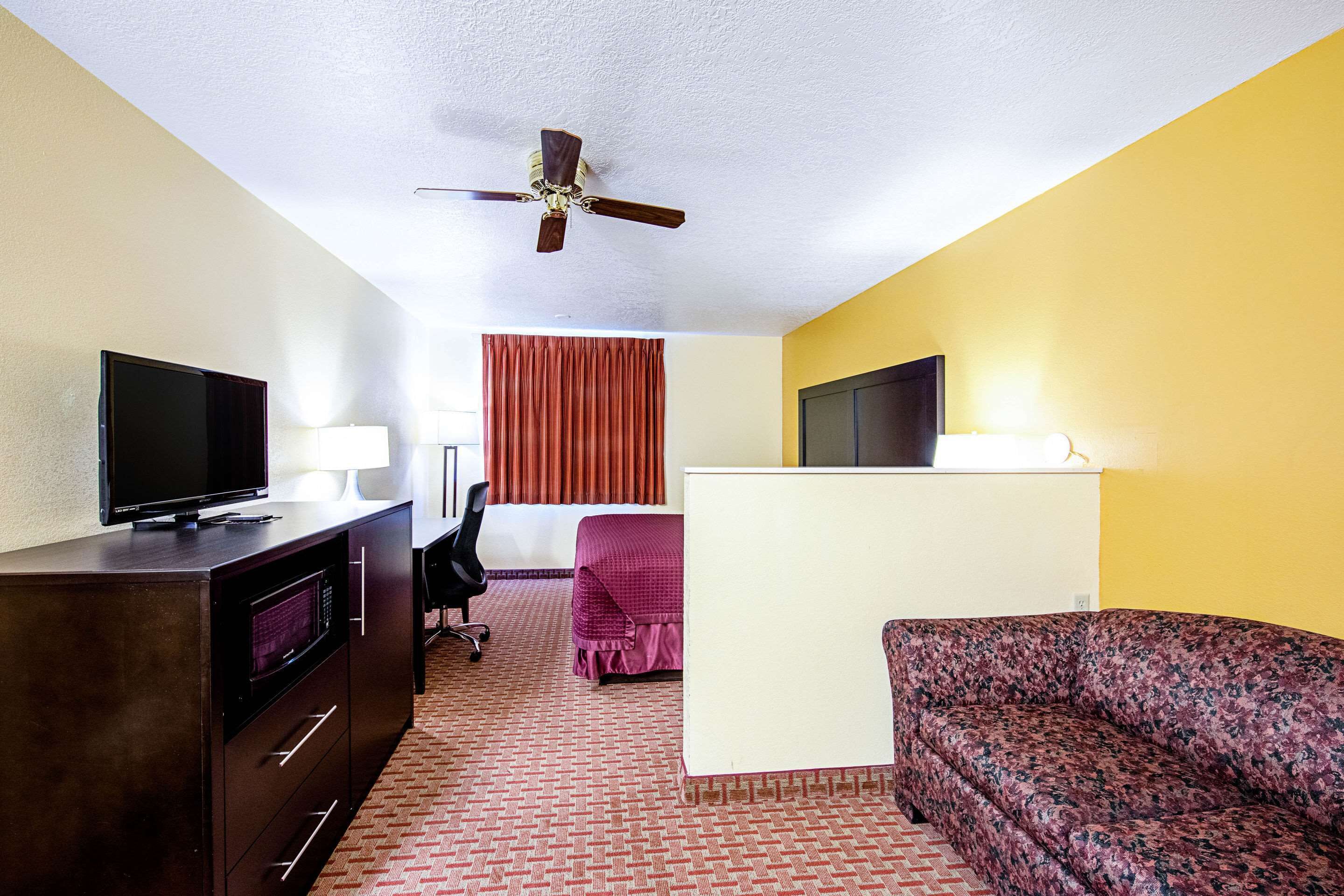 Quarto de hotel com televisão de tela plana, escrivaninha com luminária, sofá, cama com cortina vermelha e ventilador de teto.