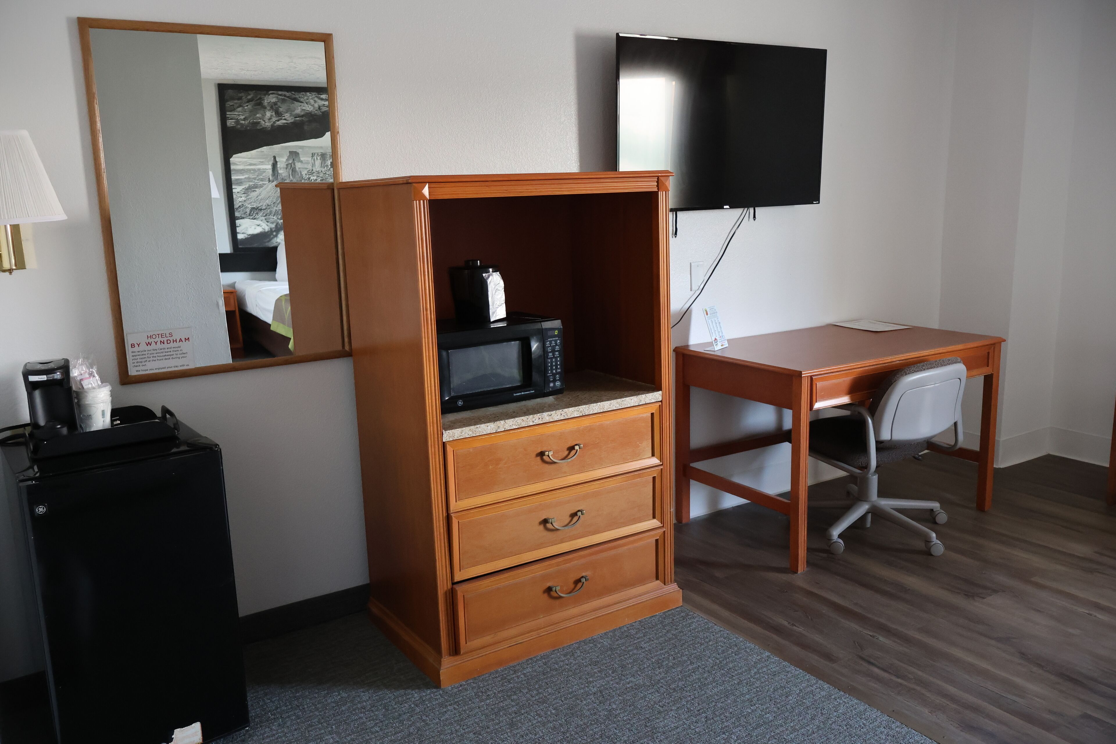 Quarto de hotel com escrivaninha de madeira, uma cadeira, um pequeno frigorífico, micro-ondas, televisão e um espelho grande.