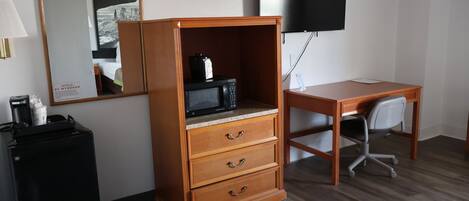 Quarto de hotel com escrivaninha de madeira, uma cadeira, um pequeno frigorífico, micro-ondas, televisão e um espelho grande.
