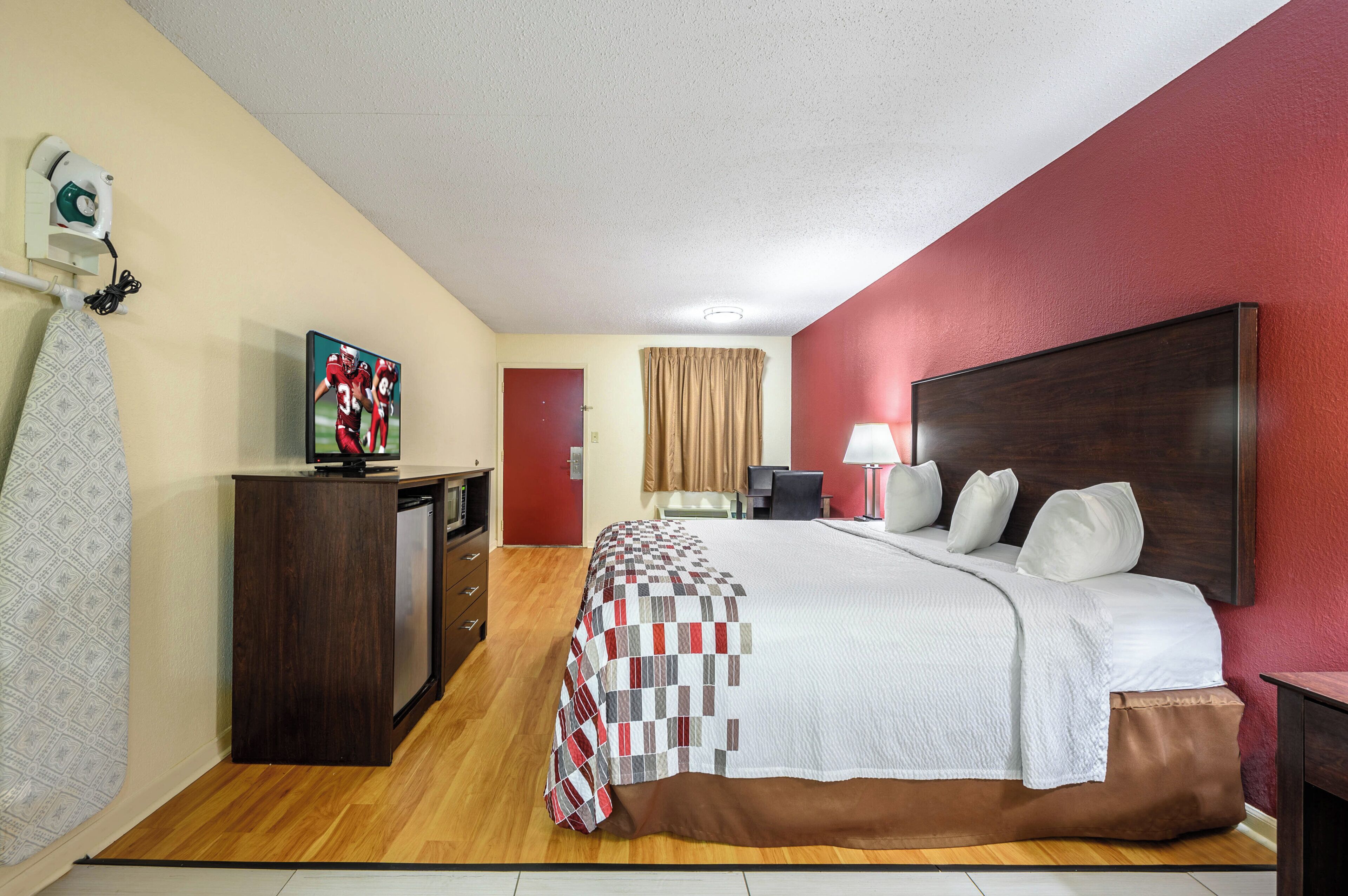 Quarto de hotel com cama, escrivaninha com televisão, cadeira, abajur e parede vermelha.