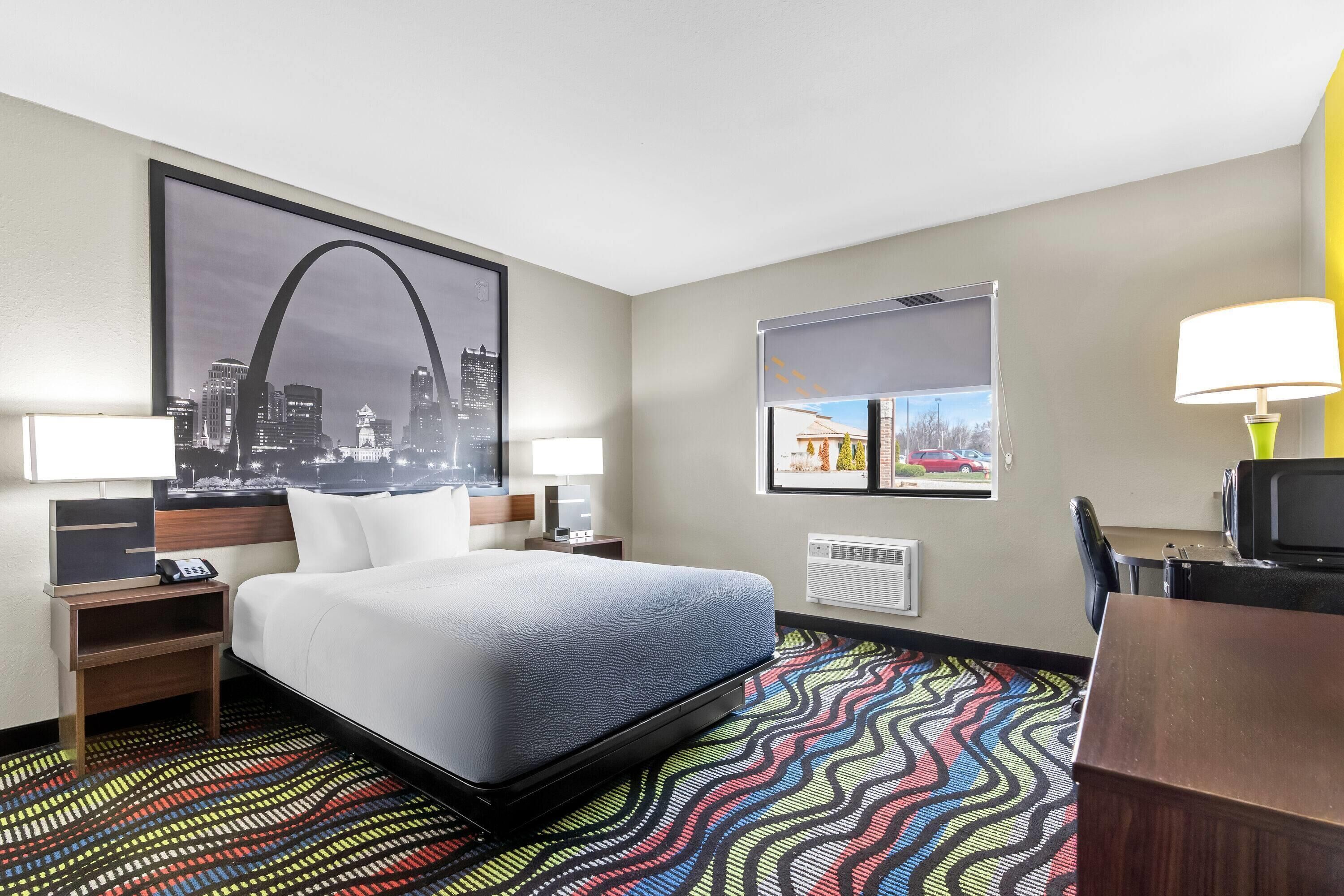 Quarto de hotel com cama, duas mesas de cabeceira, escrivaninha, cadeira, tapete colorido, pôster com vista urbana, janela e abajur.