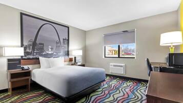 Quarto de hotel com cama, duas mesas de cabeceira, escrivaninha, cadeira, tapete colorido, pôster com vista urbana, janela e abajur.