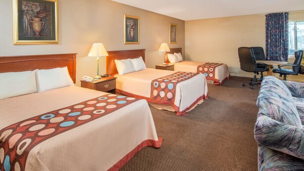 Quarto de hotel com três camas, uma escrivaninha e uma cadeira. Há quadros nas paredes e uma janela com cortinas.