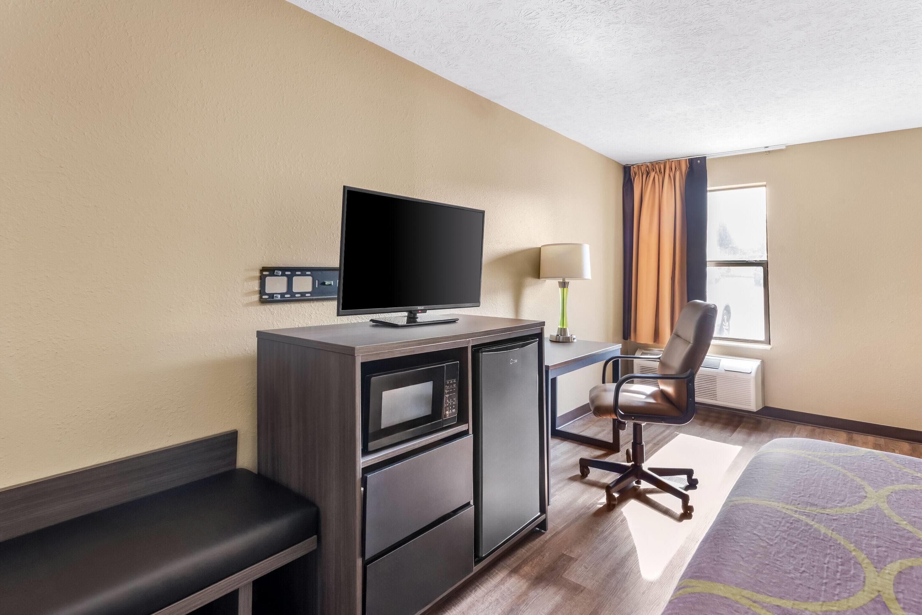 Quarto de hotel com escrivaninha, televisão de tela plana, micro-ondas, mini-geladeira, cadeira, luminária, janela com cortinas e cama com colcha roxa.