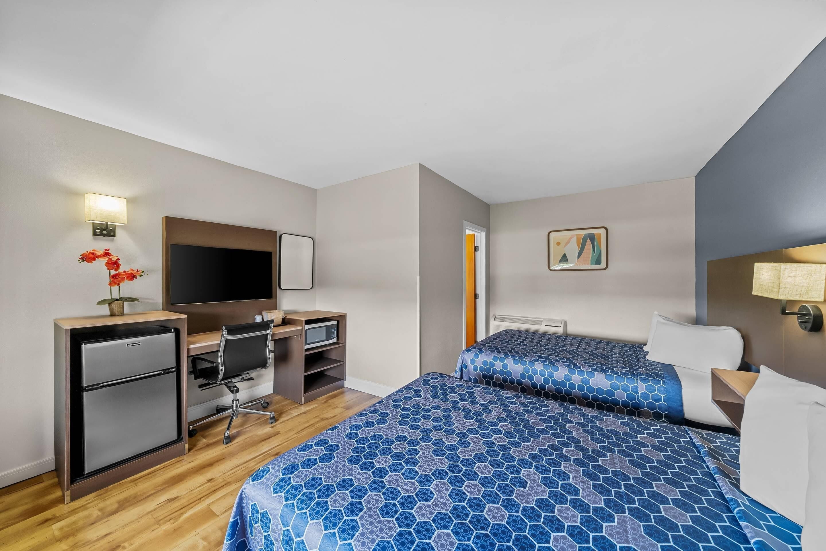 Quarto de hotel com cama, escrivaninha com computador, frigorífico, televisão e candeeiro de parede.