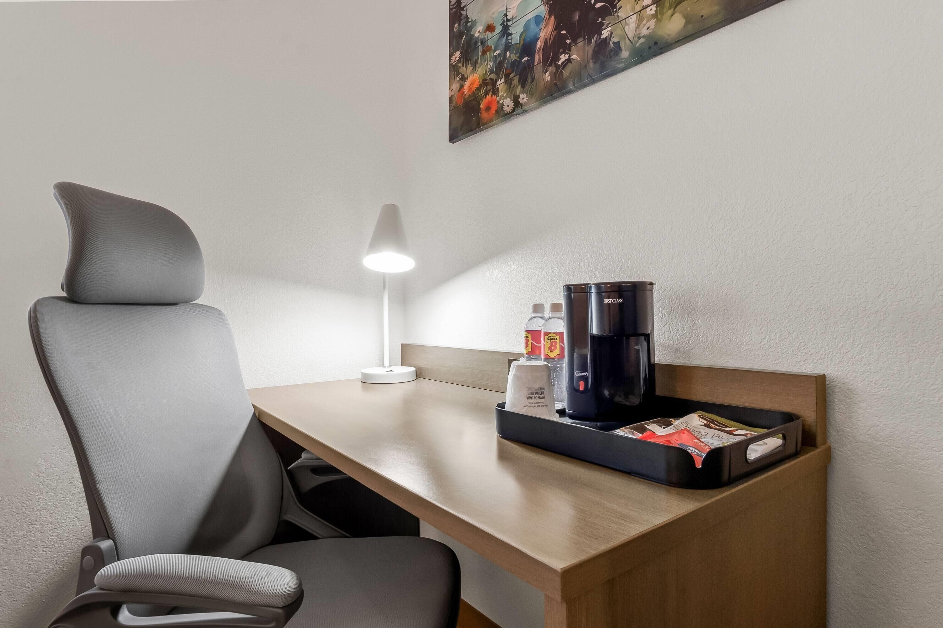 Quarto de hotel com uma escrivaninha, uma cadeira, uma luminária, uma cafeteira e um tabuleiro com objetos.