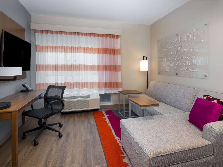 Quarto de hotel moderno com televisão de tela plana, escrivaninha com cadeira, sofá com almofada roxa, abajur e uma frase motivacional na parede.