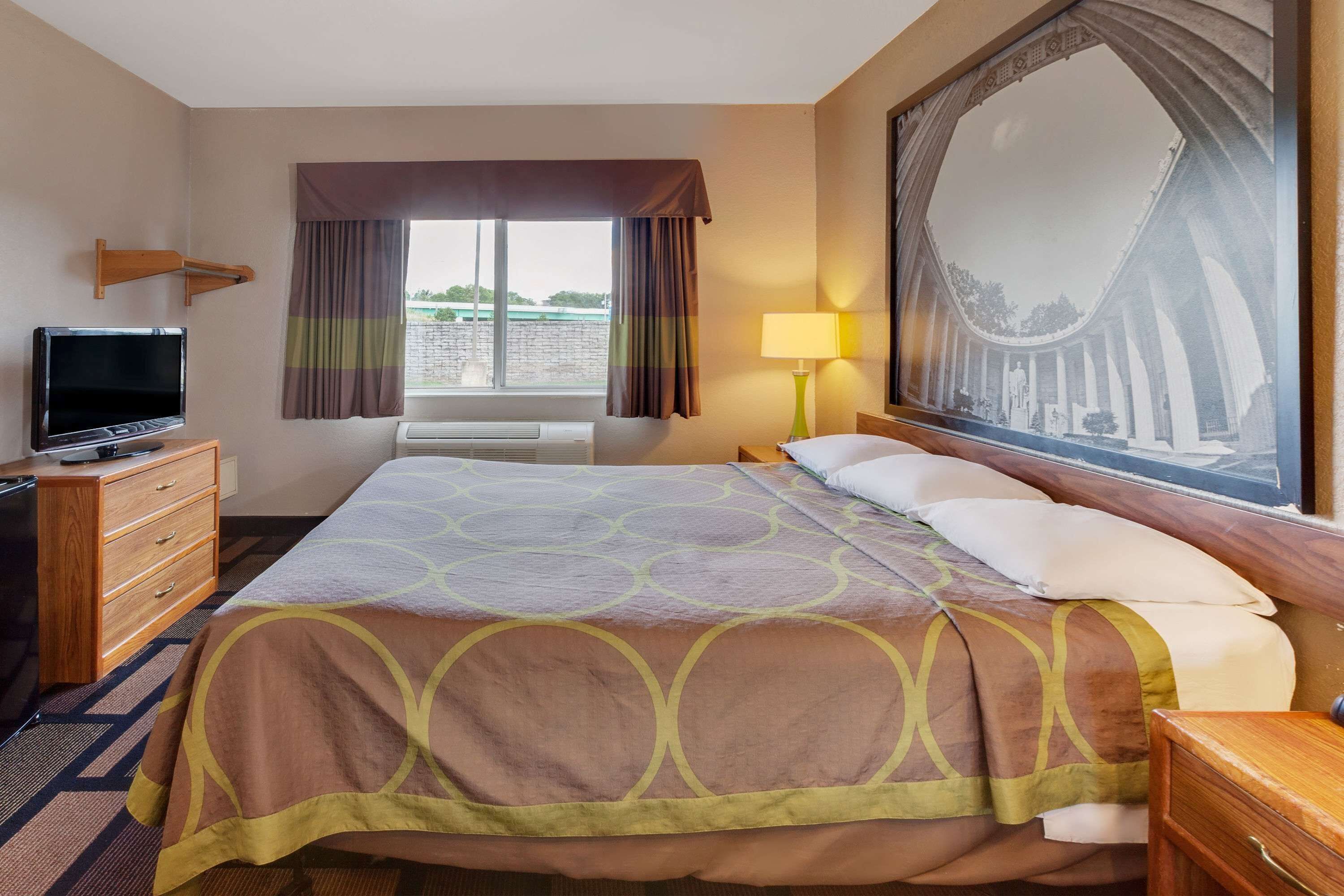 Quarto de hotel com uma cama, televisão, janela com cortinas, luminária e uma grande moldura com uma imagem na parede.