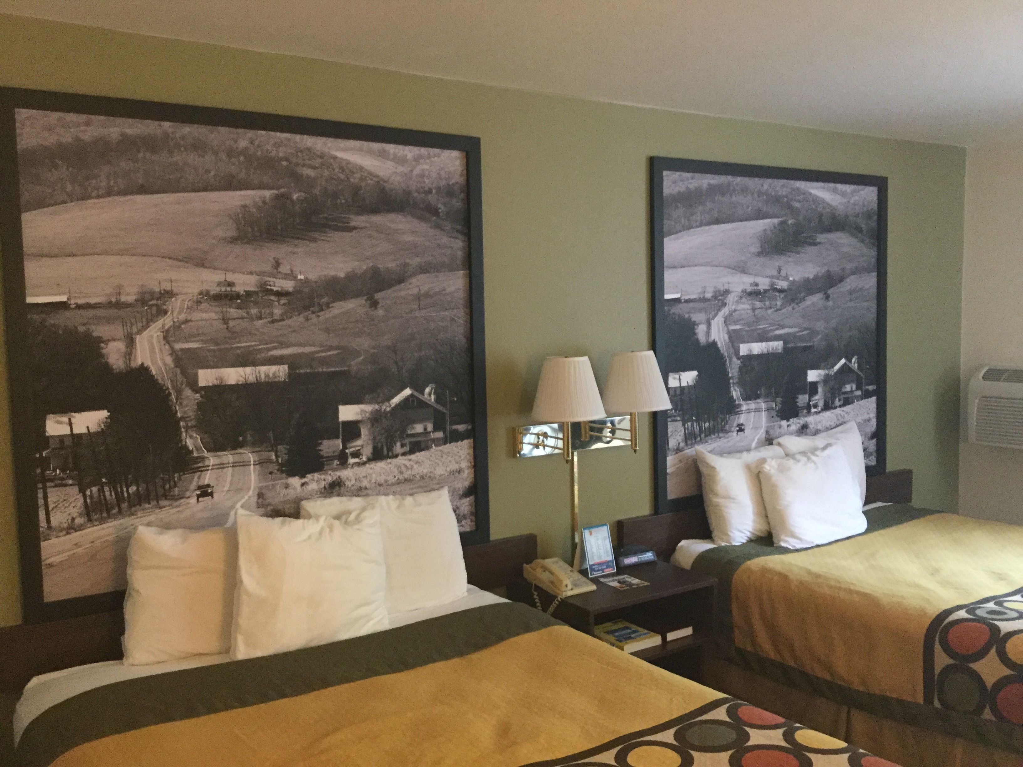 Quarto de hotel com duas camas, uma mesa de cabeceira com abajur, telefone e uma pintura em preto e branco de paisagem emoldurada na parede.
