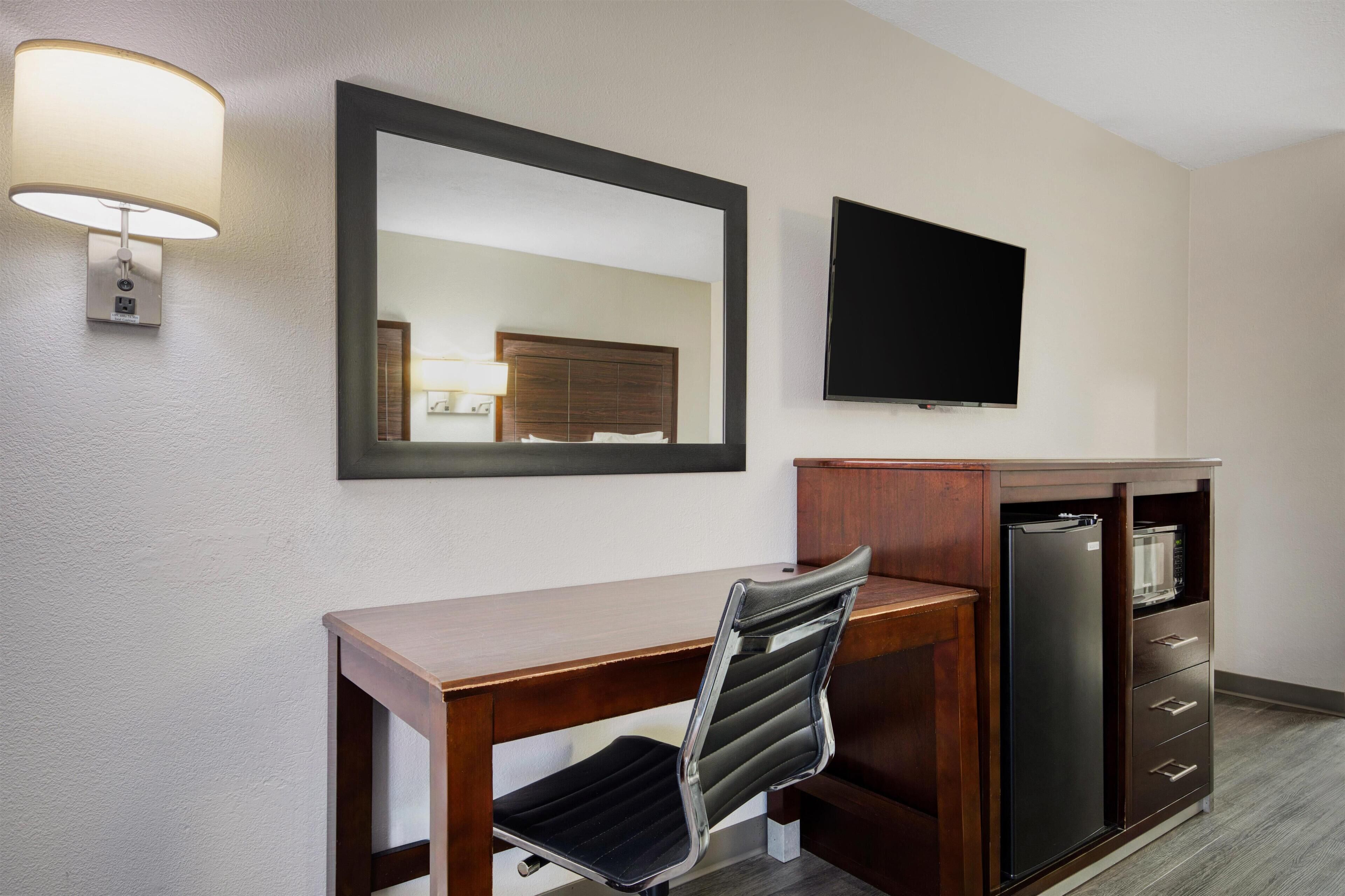 Quarto de hotel com escrivaninha de madeira, cadeira preta, televisão de tela plana, geladeira embutida e micro-ondas.