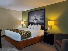 Quarto de hotel com uma cama grande, duas mesas de cabeceira com luminárias, uma poltrona e uma obra de arte na parede retratando um avião.