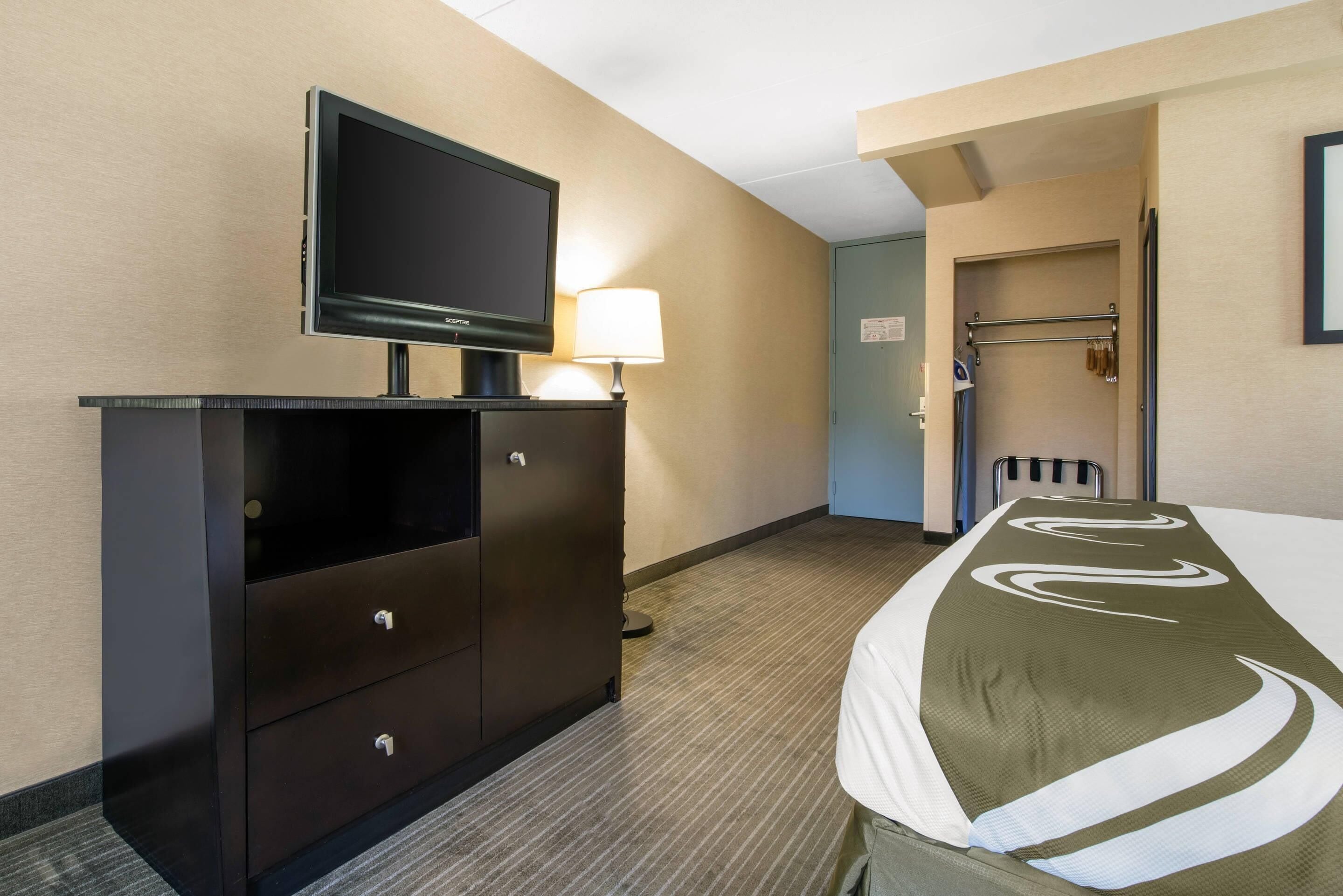 Quarto de hotel com televisão de tela plana, escrivaninha com gavetas, luminária, cama com colcha estampada, porta e cabideiro.