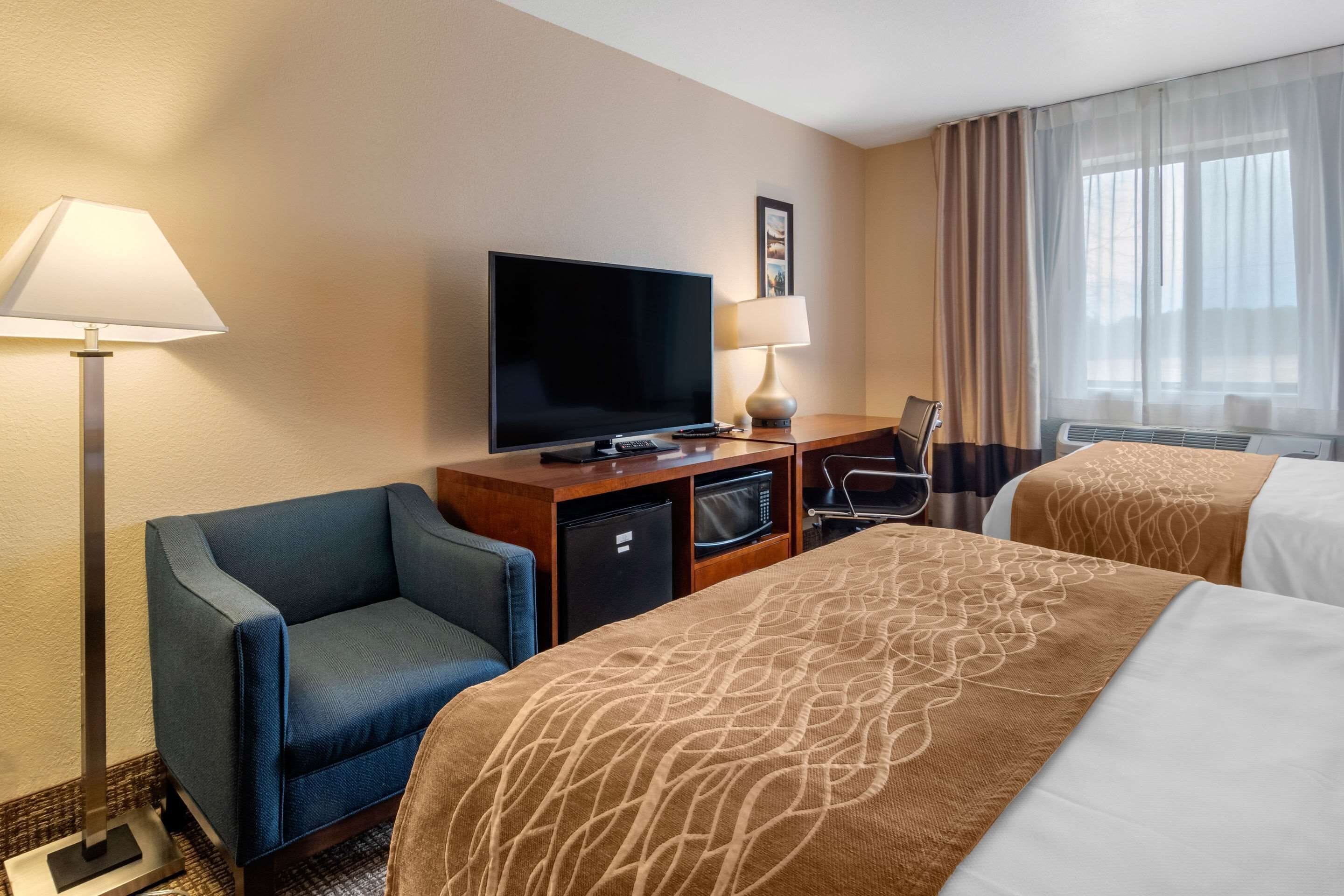 Quarto de hotel com televisão de tela plana, escrivaninha com luminária, cadeira, cama com colcha estampada e janela com cortinas.