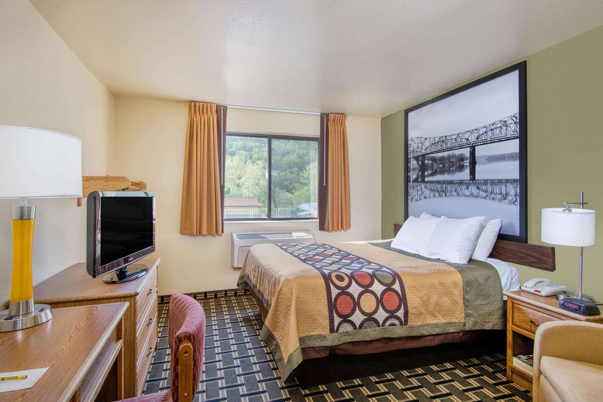 Quarto de hotel com cama, escrivaninha com televisão, cadeira, janela com cortinas e um quadro na parede.