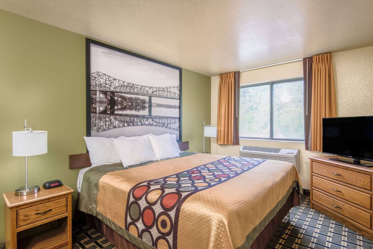 Quarto de hotel com uma cama, televisão, mesa de cabeceira, abajur e janela com cortinas.