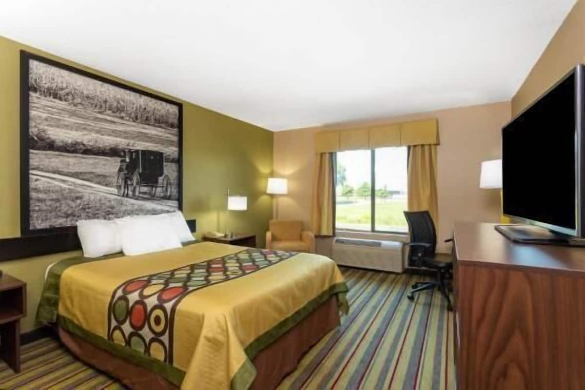 Quarto de hotel com uma cama, escrivaninha, cadeira, televisão e janela com cortinas.