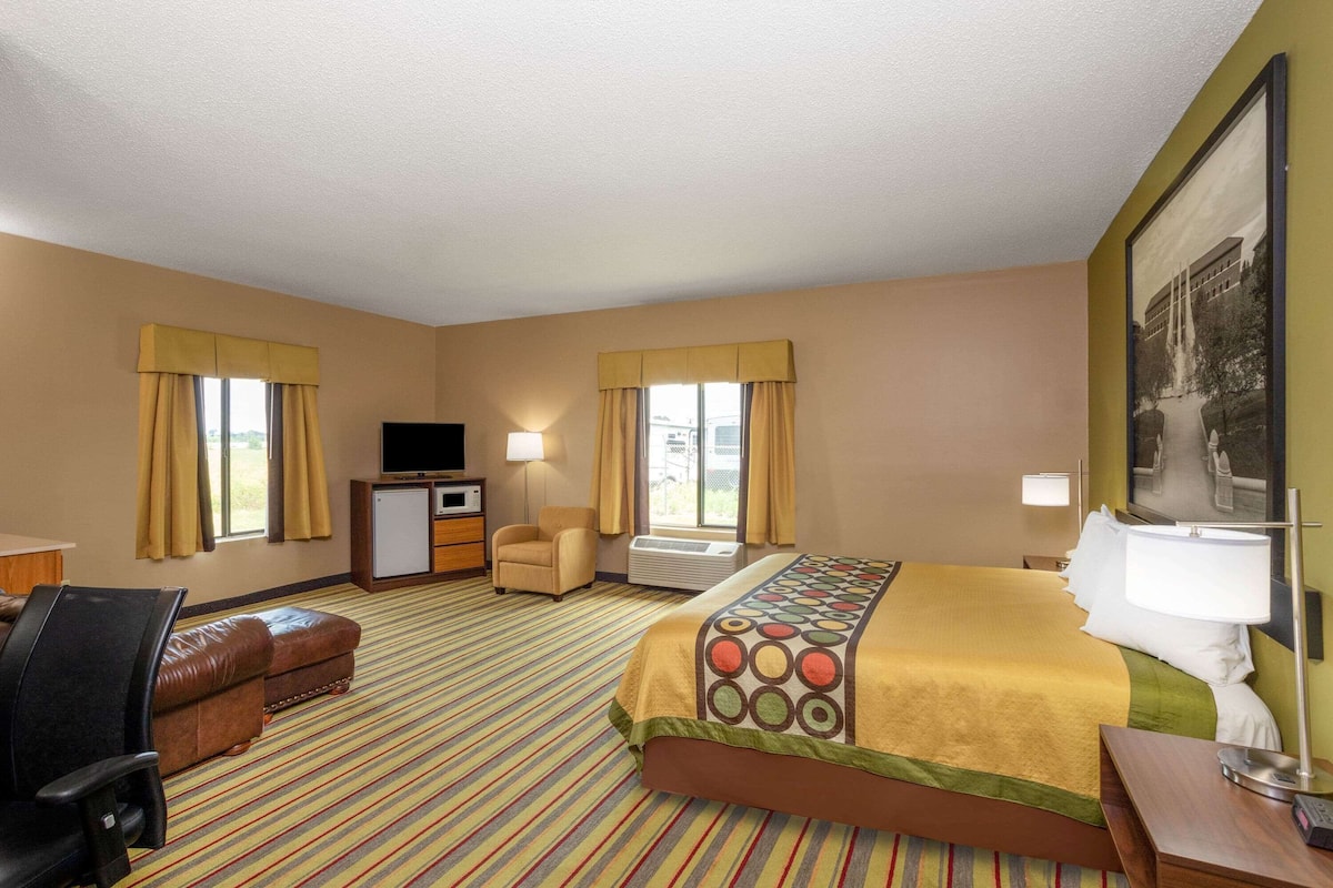 Quarto de hotel com cama, escrivaninha, poltrona, banquinho, televisão e um quadro na parede.