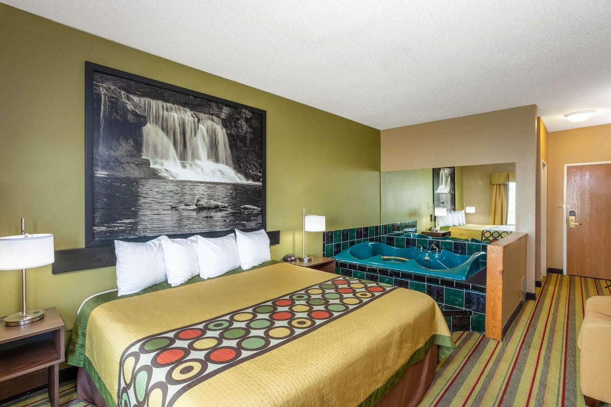 Quarto de hotel com uma cama, um quadro grande de uma cascata, casa de banho com banheira e um tapete riscado.