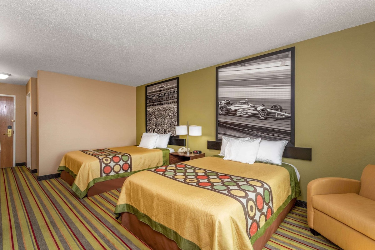 Quarto de hotel com duas camas, um sofá, um tapete listrado e uma janela grande com a imagem de um carro.