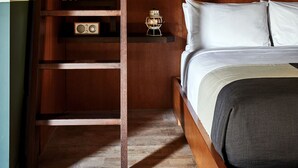 Uma cama beliche com cabeceira de madeira, uma prateleira com livros e um candeeiro, e uma mesa de cabeceira com uma lâmpada.