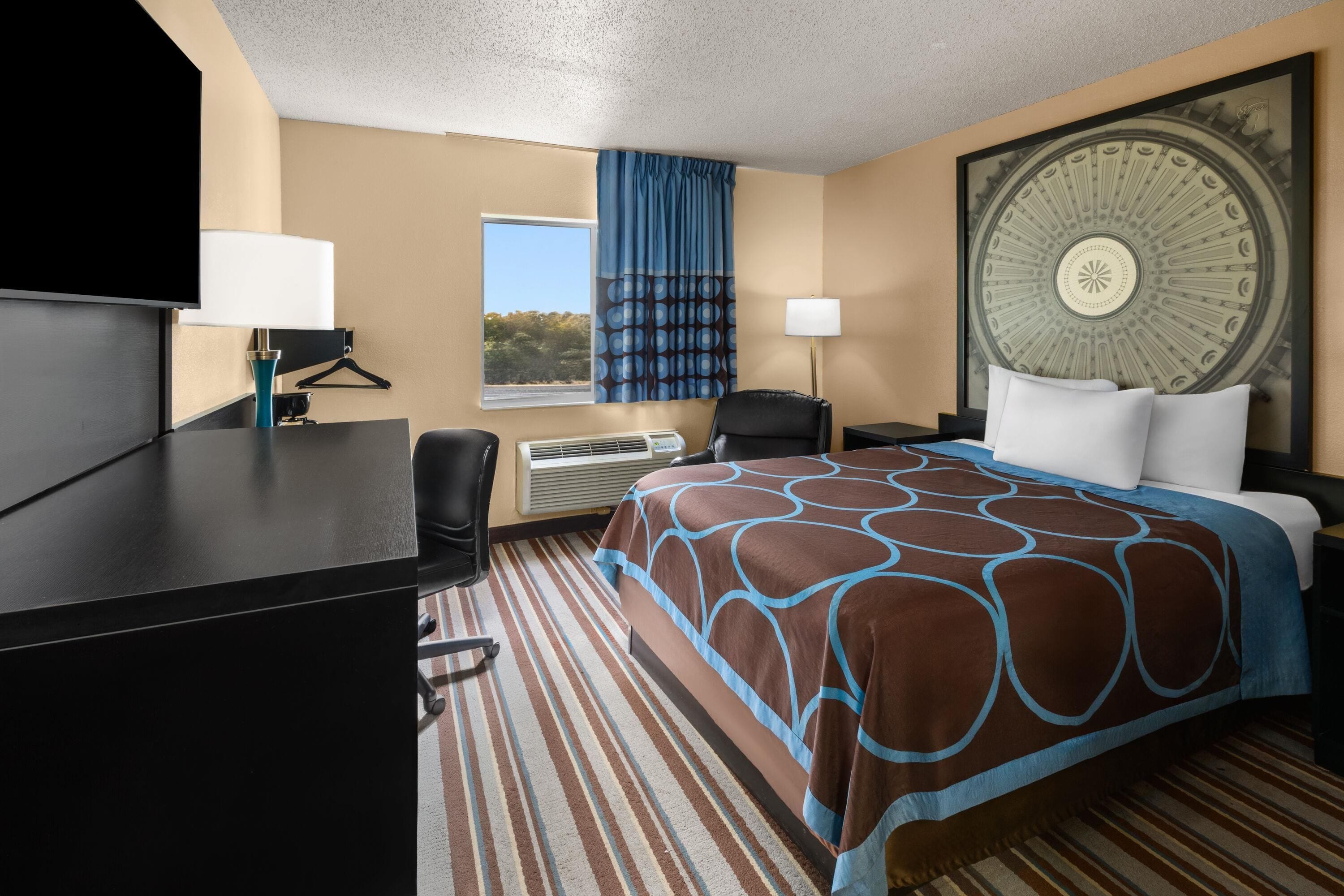 Quarto de hotel com cama, escrivaninha, cadeira, janela com cortinas, ar condicionado e relógio fixado na parede.