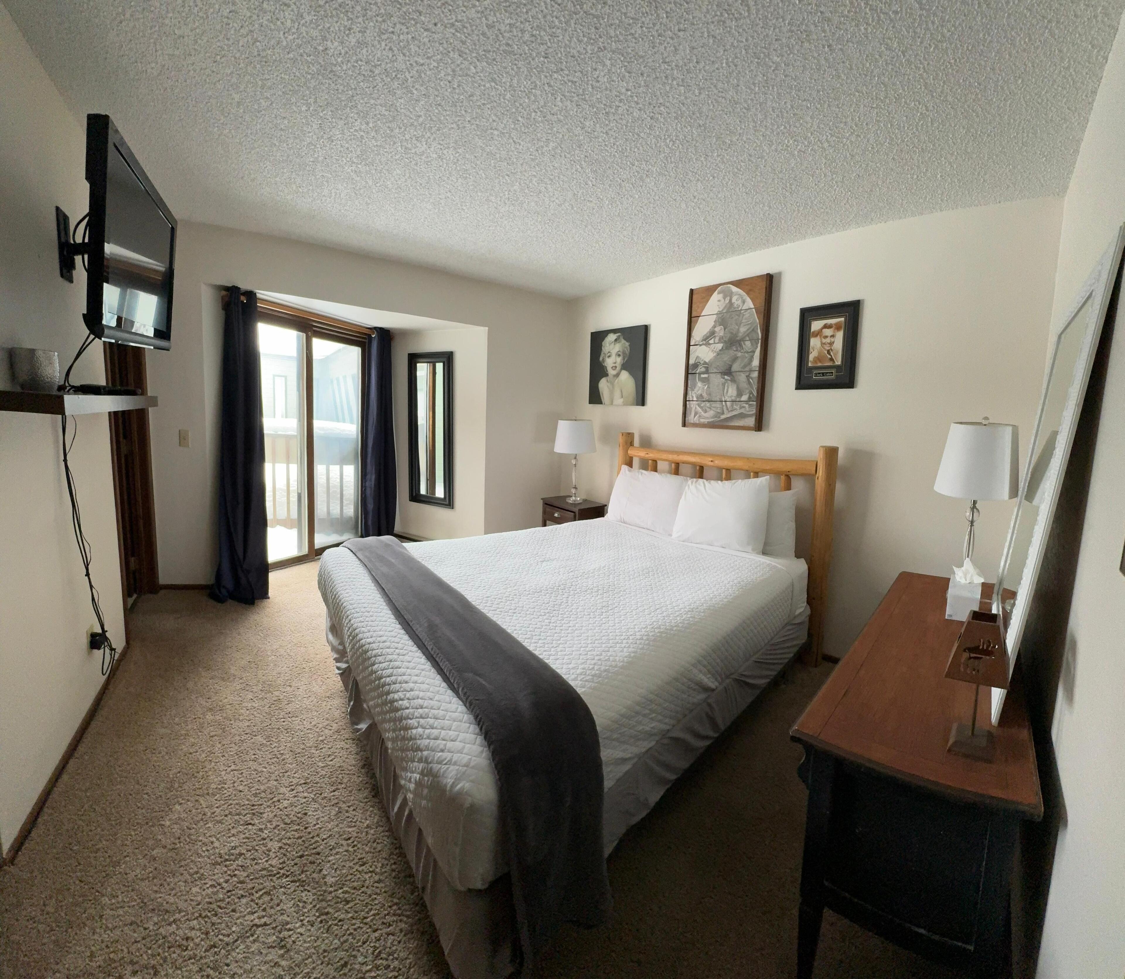Quarto de hotel com cama, televisão na parede, mesa de cabeceira com abajur e janela com cortinas.