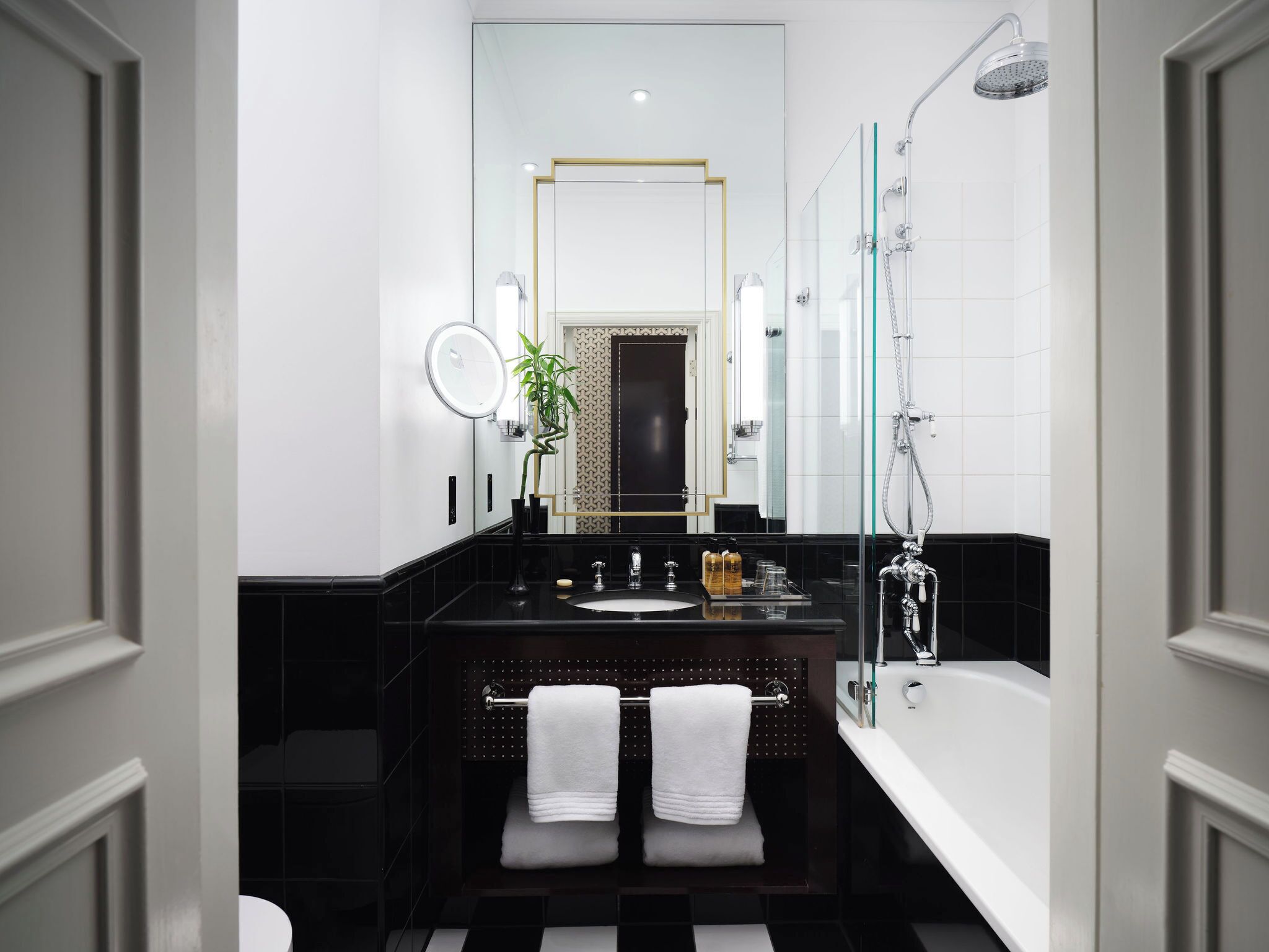 Um banheiro moderno com piso quadriculado em preto e branco, um espelho grande, uma pia com moldura dourada, um chuveiro com porta de vidro e duas toalhas brancas penduradas em um suporte.