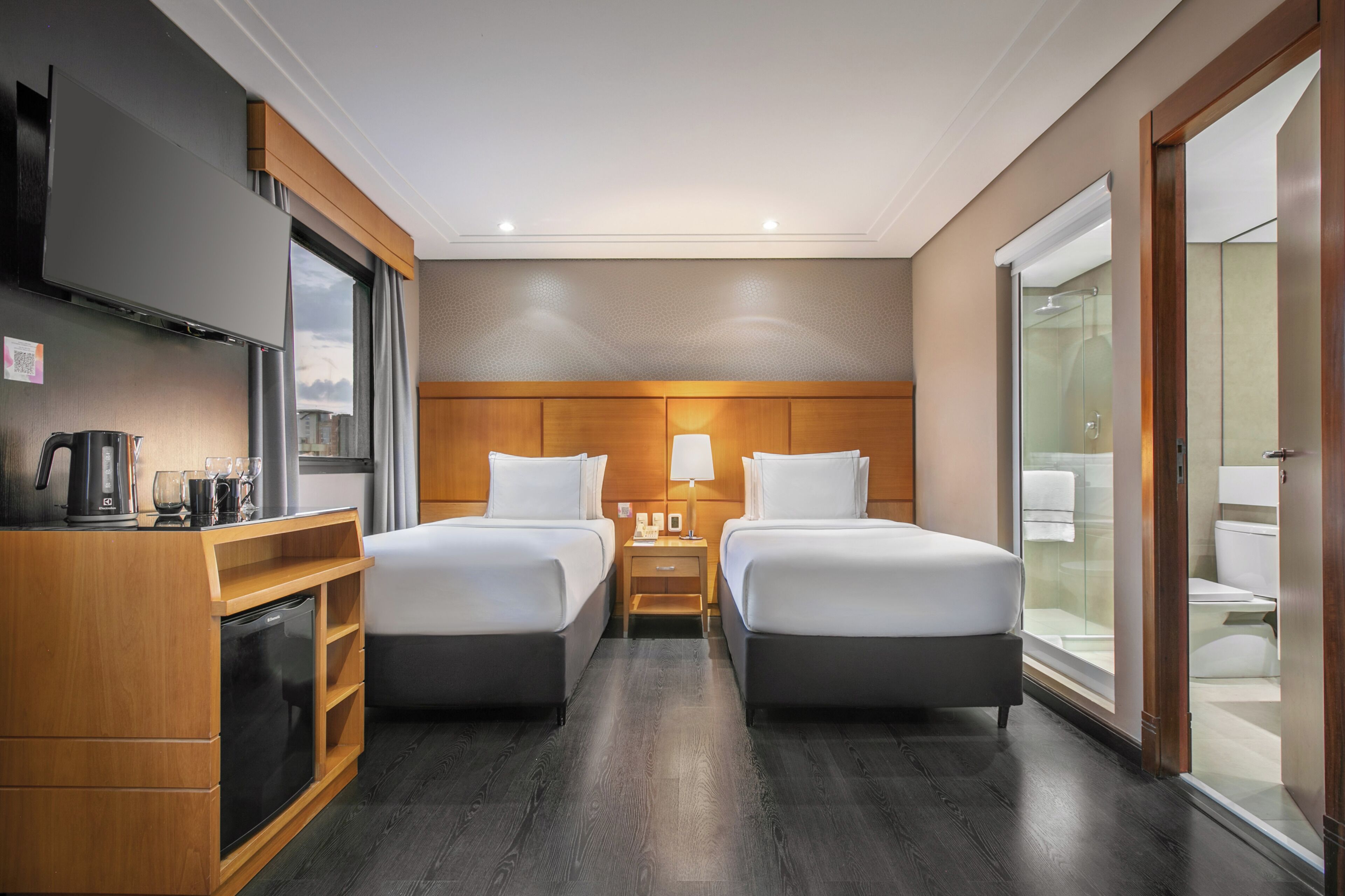 Quarto de hotel com duas camas, televisão, cafeteira, minibar, mesa de cabeceira com abajur e banheiro com box de vidro.