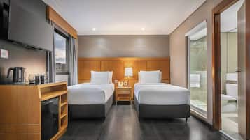 Quarto de hotel com duas camas, televisão, cafeteira, minibar, mesa de cabeceira com abajur e banheiro com box de vidro.