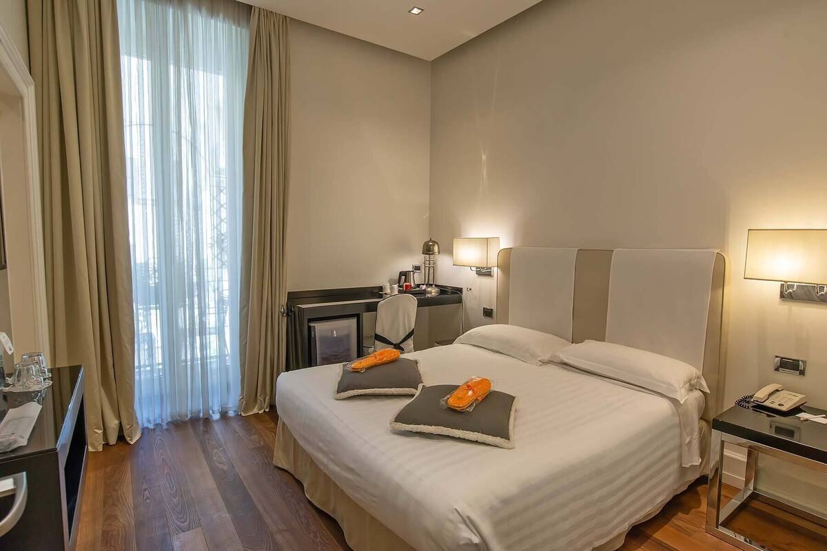 Quarto de hotel com uma cama grande, uma escrivaninha e uma cadeira. Há uma janela com cortinas, um abajur e um telefone na mesinha de cabeceira.