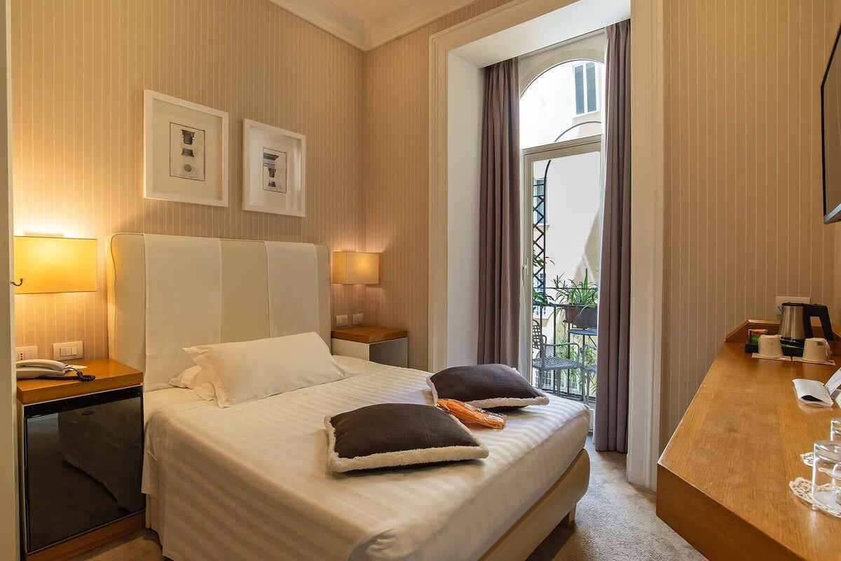 Quarto de hotel com uma cama, abajures, uma mesinha de cabeceira de madeira, uma pequena mesa com uma cafeteira e vista para a varanda.