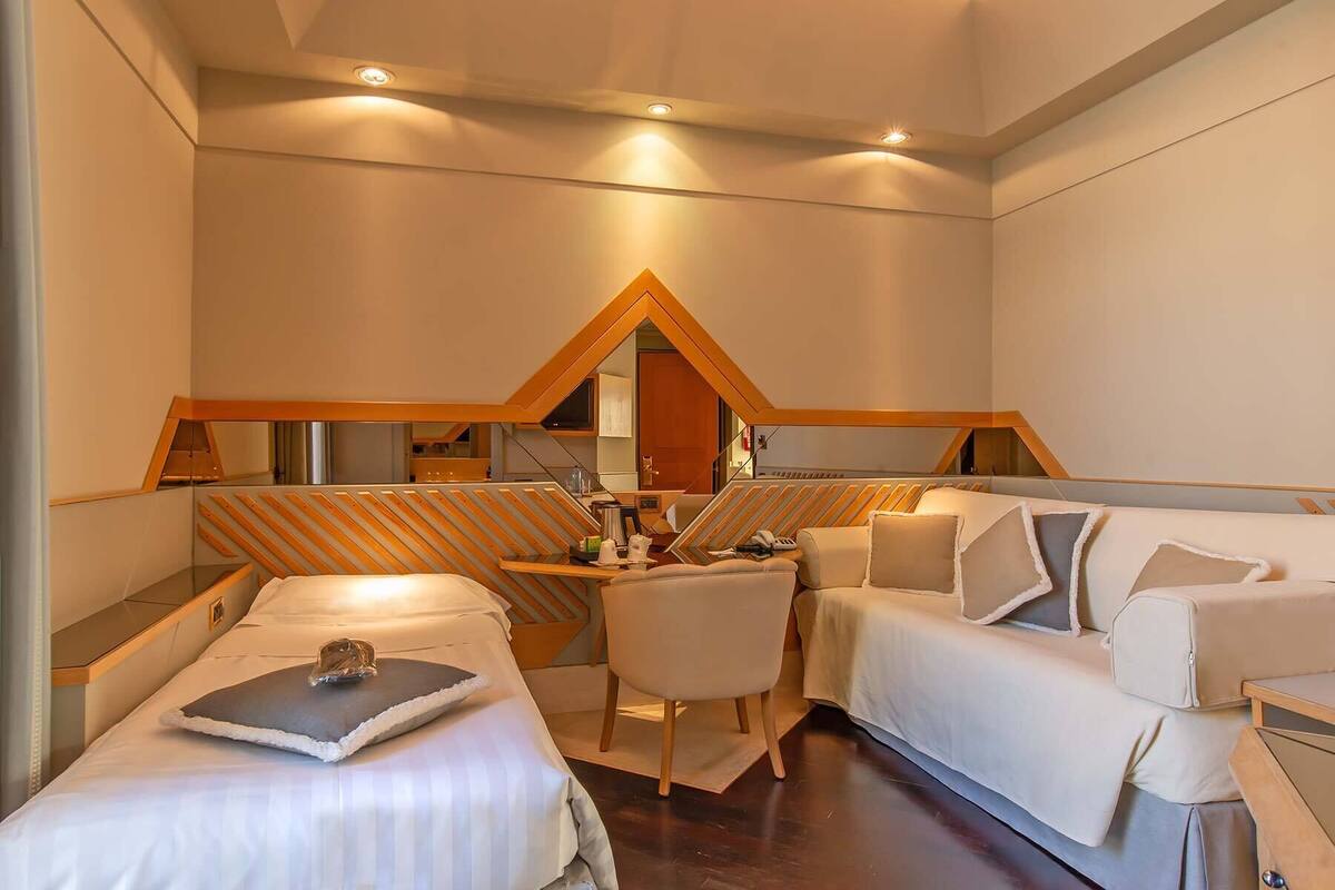 Quarto de hotel moderno com cama central, um sofá, uma escrivaninha e uma cadeira.