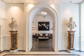 Um hall imponente com piso de mármore, um arco central e dois bustos em pedestais. Um sofá moderno e um lustre são visíveis ao fundo.