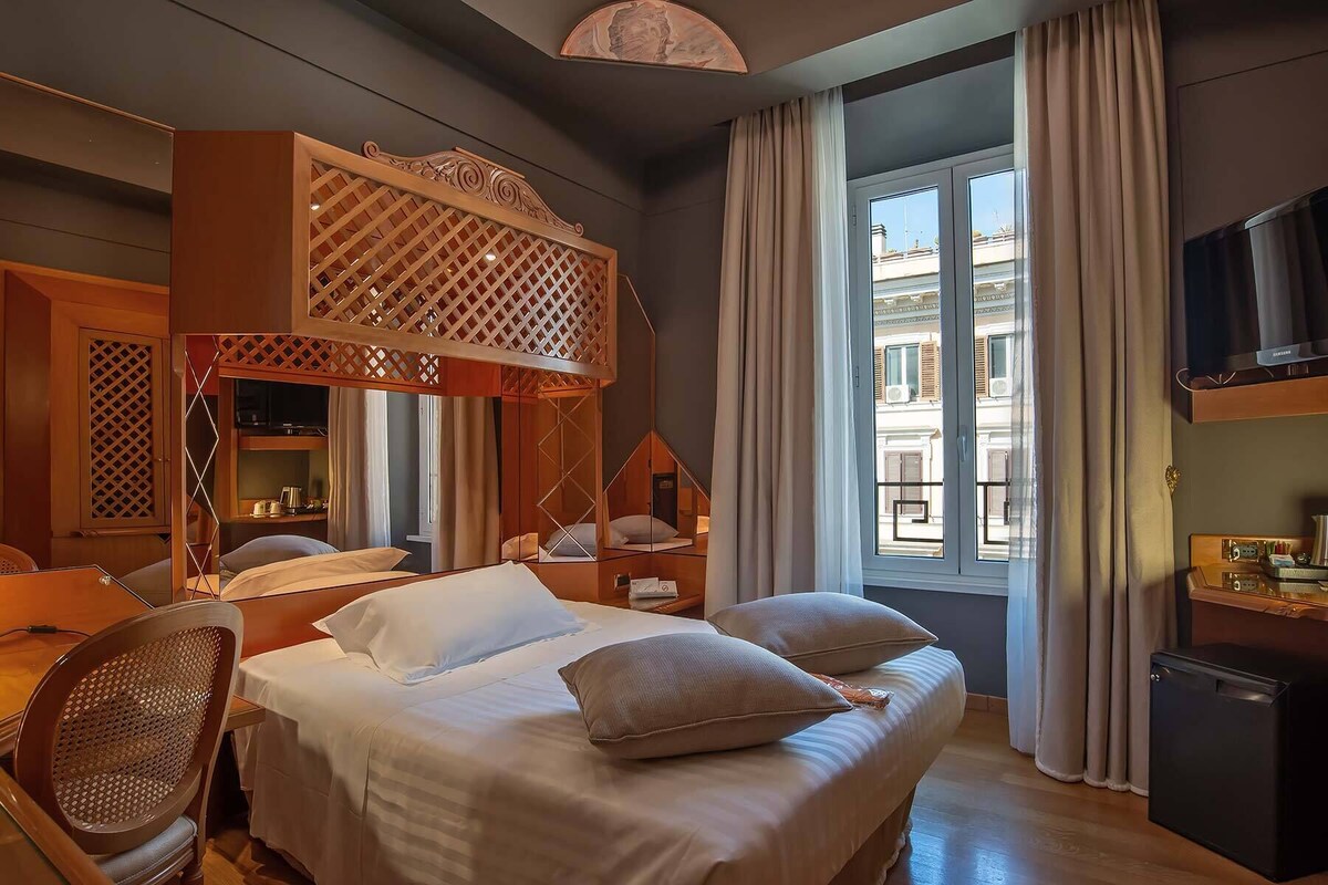 Quarto de hotel com uma cama, escrivaninha, cadeira, televisão e janela com cortinas.
