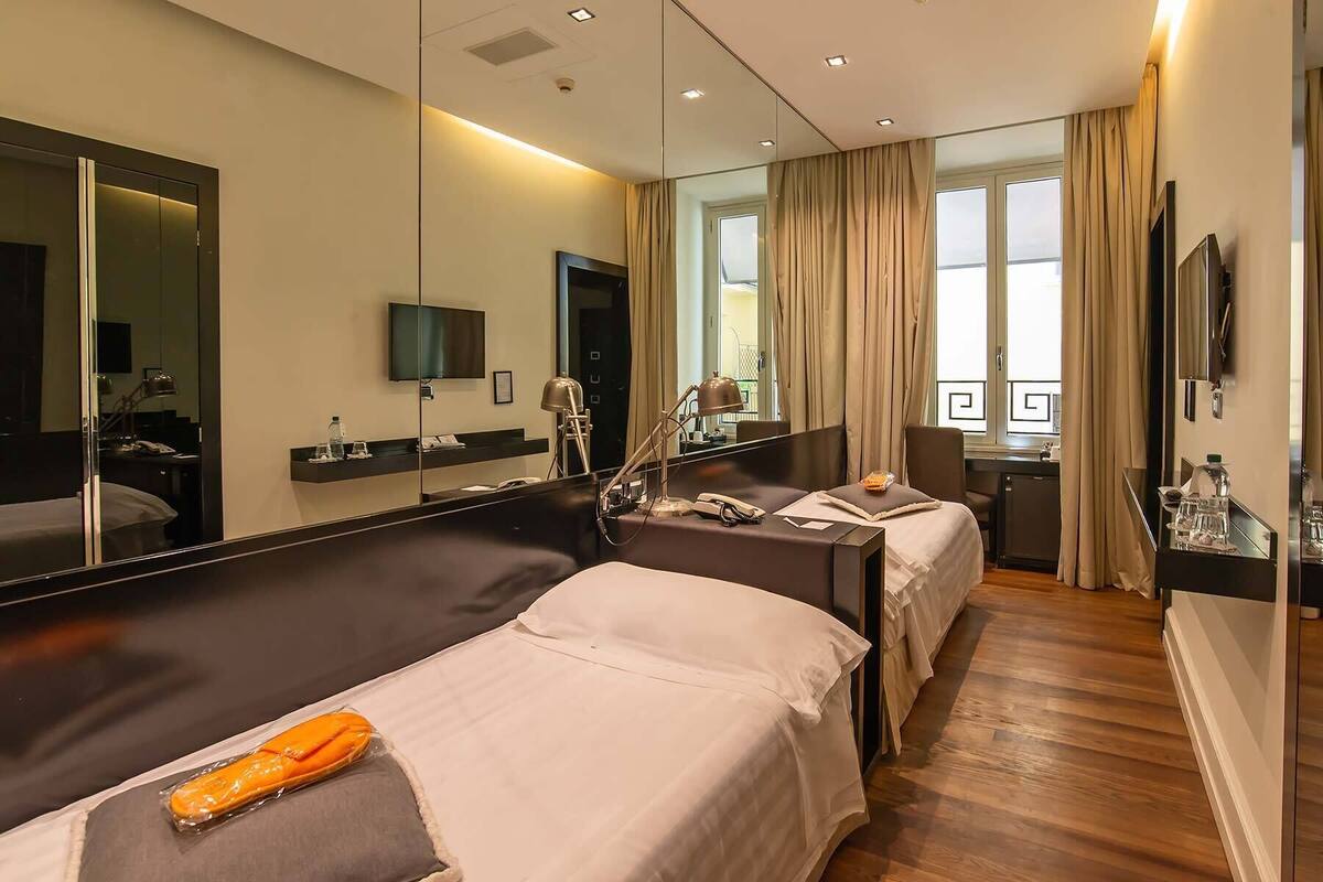 Quarto de hotel com uma cama grande, escrivaninha, cadeira, televisão e janela com cortinas.