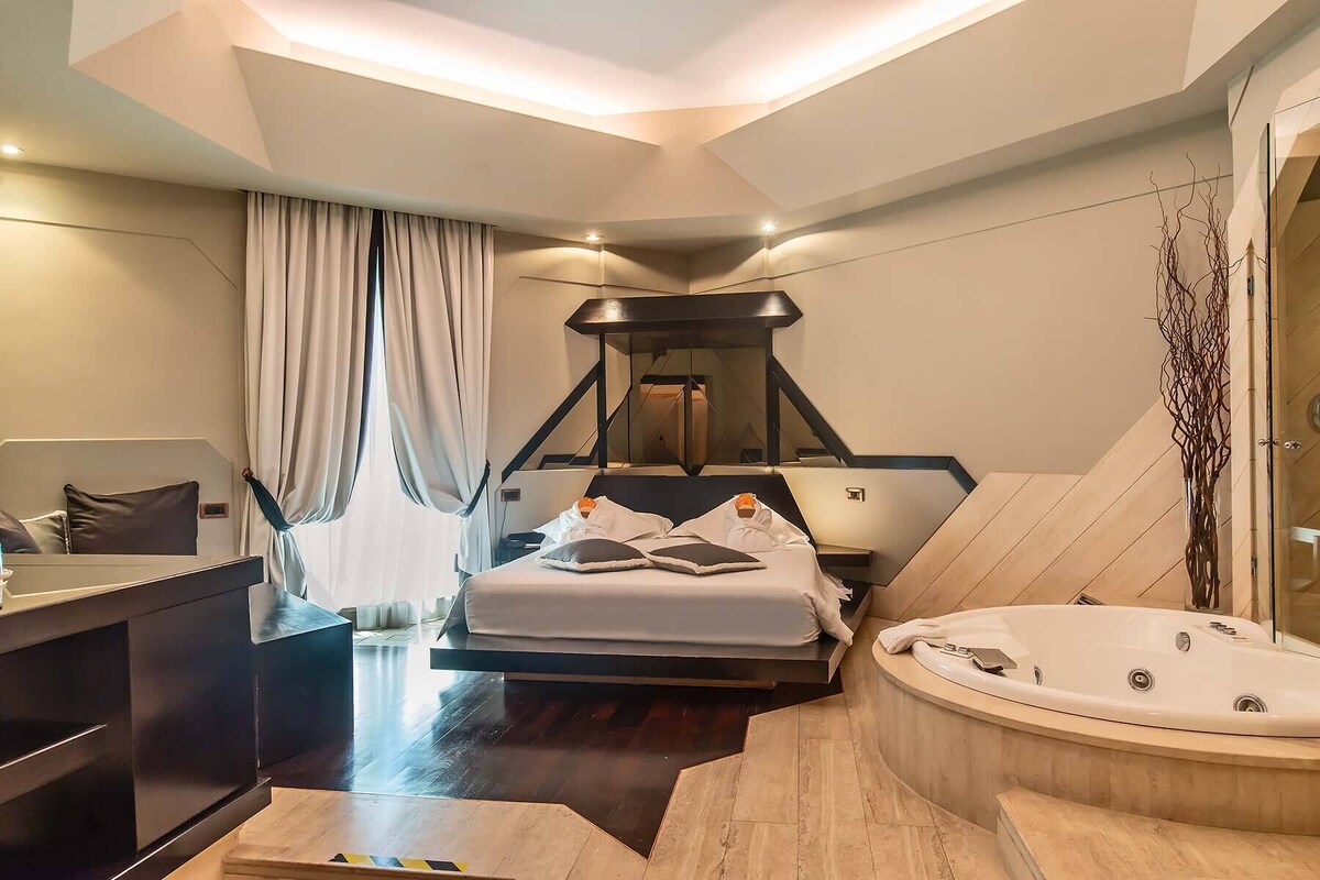 Quarto de hotel moderno com uma cama grande, uma escrivaninha, uma cadeira, luminária de mesa, um espelho amplo, uma banheira e uma janela com cortinas.
