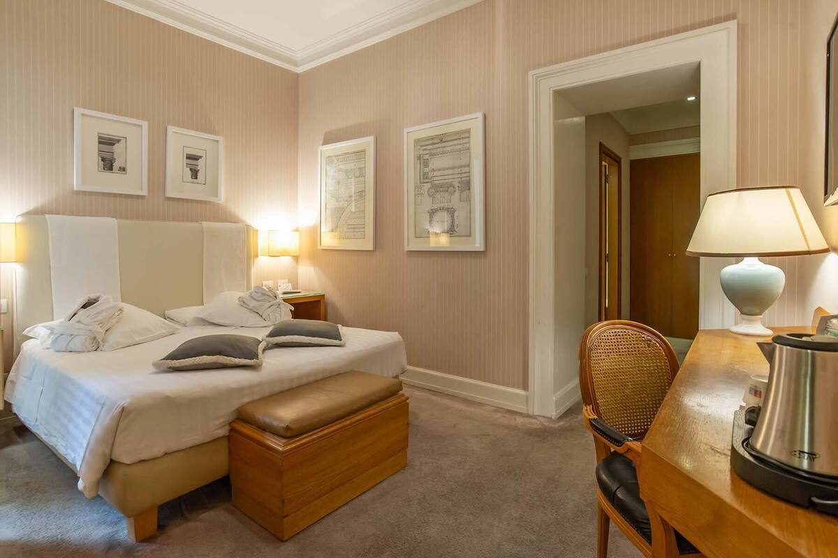 Quarto de hotel com cama, um banco de madeira, escrivaninha com abajur e quadros na parede.