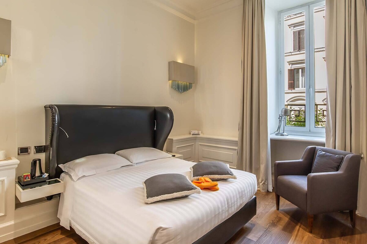 Quarto de hotel com uma cama grande, mesa de cabeceira, uma cadeira e janela com cortinas.