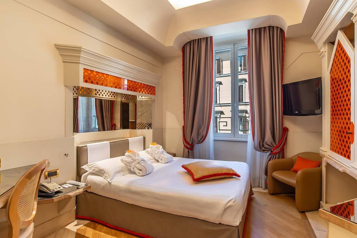 Quarto de hotel com cama, escrivaninha, cadeira, televisão e janela com cortinas.