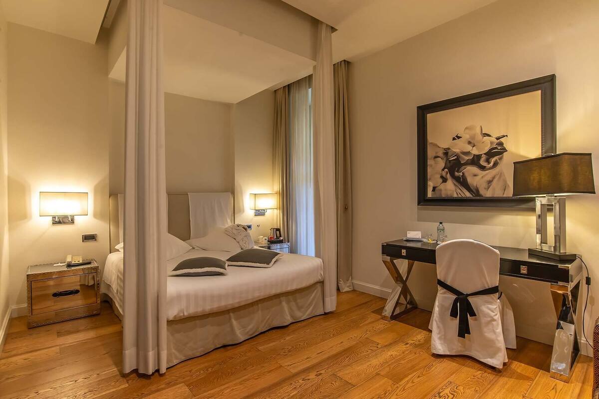 Quarto de hotel com uma cama de dossel, uma escrivaninha com cadeira, uma mesinha de cabeceira e um quadro na parede.