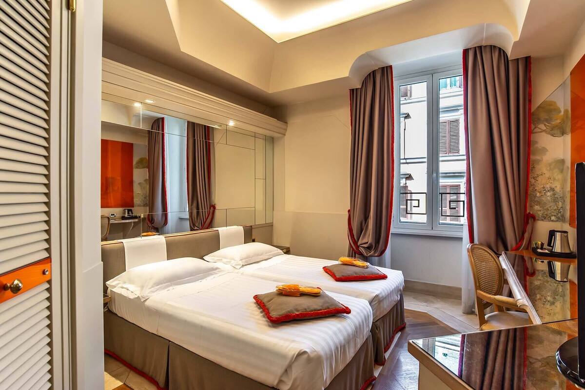 Quarto de hotel com duas camas, um espelho grande, uma janela com cortinas e uma pequena mesa com uma cadeira.