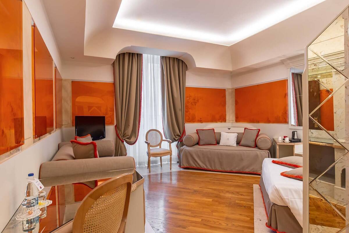 Quarto de hotel moderno com sofá, cama, cadeira e uma pequena mesa com garrafa e copos.