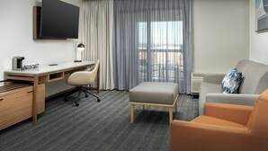 Quarto de hotel com televisão de tela plana, escrivaninha com cadeira, sofá e janela com cortinas.