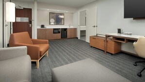 Quarto de hotel moderno com sofá, escrivaninha, cozinha compacta e televisão de tela plana.