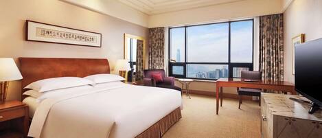 Quarto de hotel com uma cama grande, uma escrivaninha, uma cadeira, televisão e vista para o horizonte da cidade.