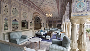Um quarto luxuoso com teto e paredes ricamente decorados, uma área de estar com sofás e pufes, e uma janela ampla.
