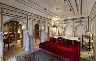 Um quarto luxuoso com uma cama grande, arcos ornamentados e um espaço de estar com um sofá vermelho e uma mesa.