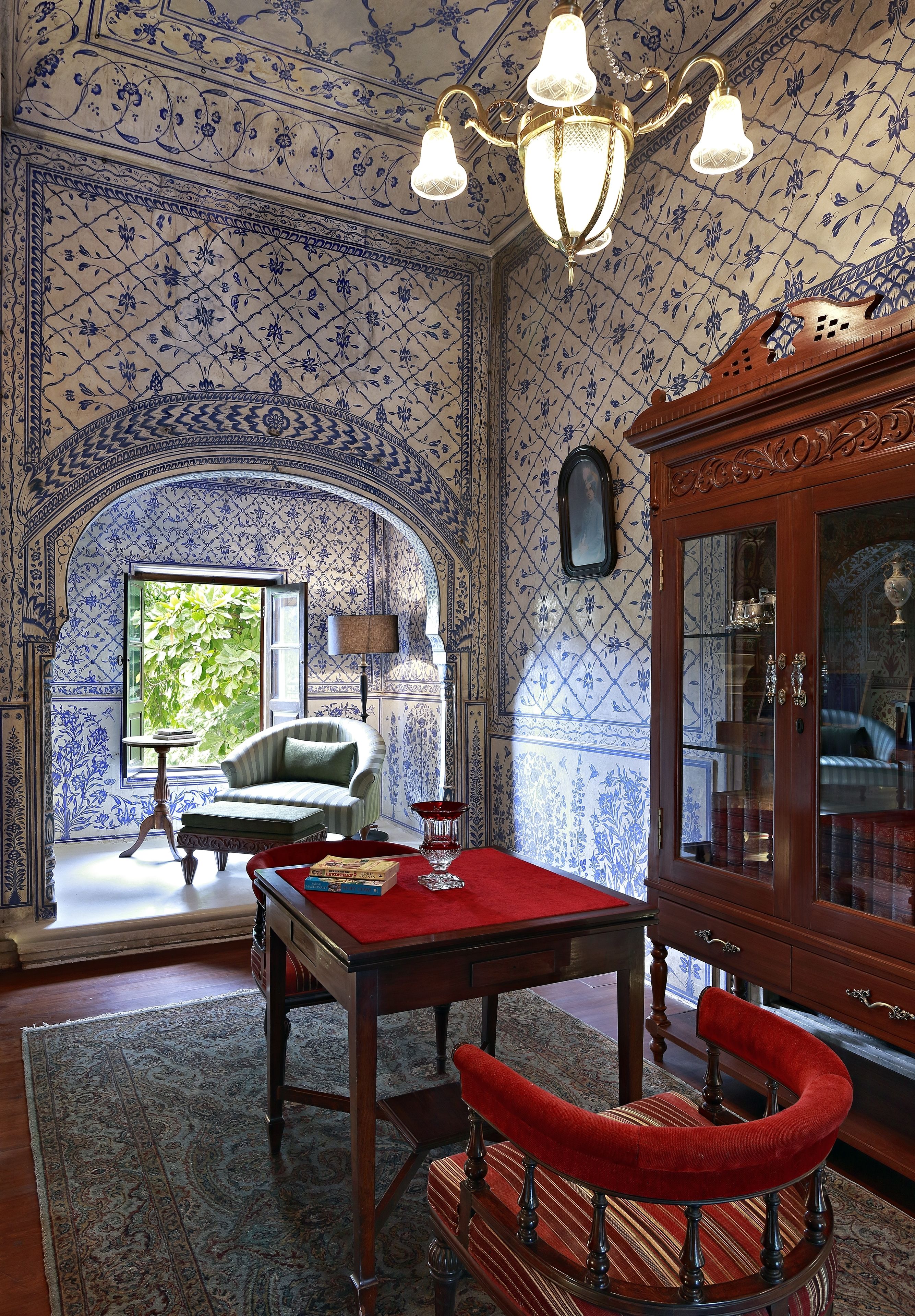 Um quarto com parede revestida de azulejos azuis e brancos estampados, um armário de madeira, uma cadeira com assento vermelho, uma pequena mesa com toalha vermelha e uma janela com vista para o verde.
